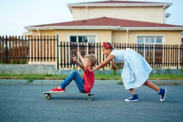 Glückliches Mädchen, das Skateboard mit ihrem Bruder entlang Zaun des neuen Hauses drückt